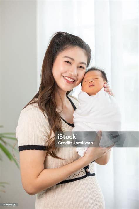 아늑한 집의 창문 앞에 서있는 갓 태어난 아기를 품에 안고있는 아름다운 아시아 여성 행복한 유아 아기는 어머니 팔에 안전하고
