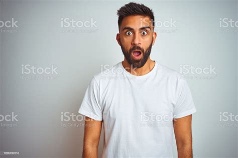 고립 된 흰색 배경 위에 서있는 티셔츠를 입은 젊은 인도 남자는 놀라움과 놀라움의 표정 두려움과 흥분된 얼굴로 충격을 받았습니다