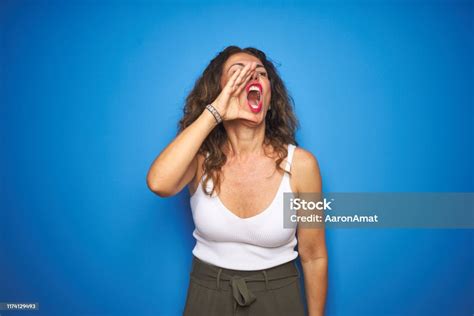 파란색 고립 된 배경 위에 서있는 곱슬 머리를 가진 중년 노인 여성은 입에 손을 대고 큰 소리로 외치고 비명을 지습니다 통신 개념입니다 비명에 대한 스톡 사진 및 기타