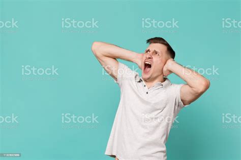 스튜디오에서 파란색 청록색 배경에 고립 된 손으로 귀를 덮고 옆으로 바라보는 캐주얼 한 옷을 입고 비명을 지르는 젊은 남자를
