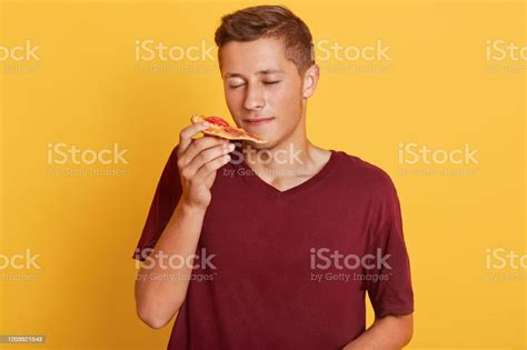 즐거운 달콤한 어린 소년의 초상화는 그의 눈을 감고 피자 조각을 들고 피자를 좋아하고 캐주얼 한 어두운 빨간 티셔츠를 입고 스튜디오에서 노란색 배경 위에 고립 되어 있습니다