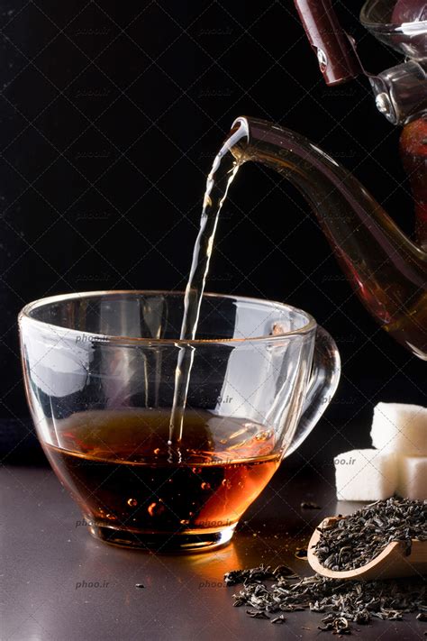 عکس ریختن چای از قوری به فنجان در کنار قاشق چوبی با چای خشک و قند در پس