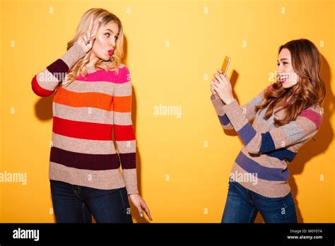 Cheery Girl Taking A Photo Of Her Girlfriend Isolated Over Yellow
