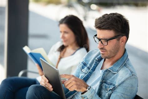 man using tablet and woman reading book at bus stop stock image image