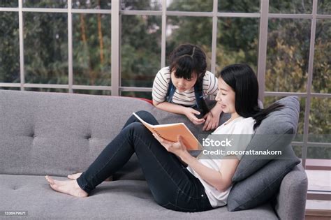 웃고 있는 아시아 인 어머니와 어린 소녀 아이가 거실에서 소파에서 함께 책을 읽고 있습니다 홈스쿨 및 교육 개념 2명에 대한