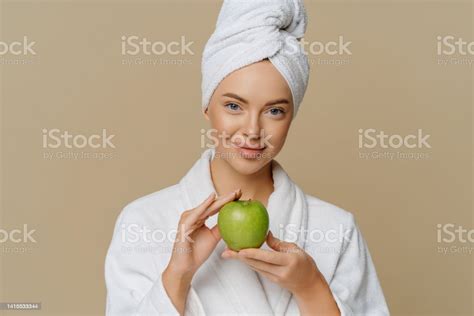 사랑스러운 젊은 여성은 머리에 드레싱 가운 을 입고 목욕을 복용 한 후 피부를 상쾌하게하고 갈색 배경에 고립 된 많은 비타민을 포함하는 녹색 사과를 보유하고 있습니다 위생
