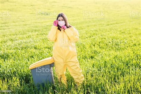 노란색 보호복을 입은 한 여성이 분홍색 장갑을 끼고 보호 마스크를 착용한 초록색 밭 한가운데에 서 있다 한 여성이 쓰레기통 근처에 서서 지구를 구하기 위해 전화합니다 25