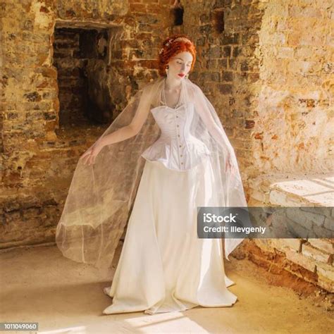 동화에서 오래 된 성곽에 있는 헤어스타일으로 젊은 르네상스 빨간 머리 공주 코르 셋에 인형 궁전에서 르네상스 공주입니다 오래 된 돌 담의 배경으로 하얀 드레스에 동화 여왕