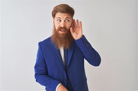 고립 된 흰색 배경 위에 서있는 양복을 입은 젊은 빨간 머리 아일랜드 사업가가 소문이나 험담에 귀를 기울이며 귀에 손을 대고 미소짓고 있습니다 청각 장애 개념입니다 경영자에