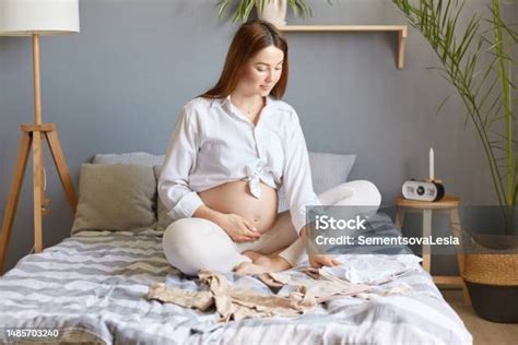 흰 옷을 입고 배꼽이 드러난 매력적인 행복한 백인 임산부는 밝은 침실에서 침대에 앉아 아기를 위한 작은 옷을 고르고 출산을