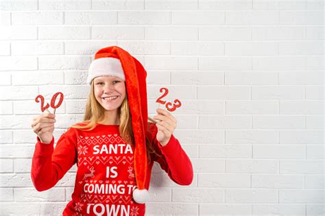 빨간 스웨터와 산타 모자를 쓴 어린 소녀가 텍스트를 위한 장소가 있는 흰색 벽돌 벽에 2023이라는 숫자를 손에 들고 있습니다 2023년에 대한 스톡 사진 및 기타 이미지