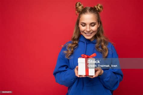 파란 트렌디한 후드를 입고 빨간 리본으로 흰색 상자를 바라보는 빨간 배경 벽에 고립 된 매력적인 행복한 미소 젊은 금발 여성의 샷 경외감에 대한 스톡 사진 및 기타 이미지