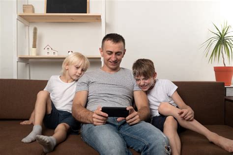 Dad And Two Sons Are Sitting On Couch And Watching A Video On Phone Happy Family Stock Photo