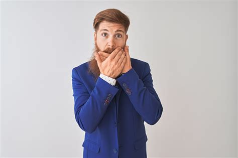 고립 된 흰색 배경 위에 서있는 양복을 입은 젊은 빨간 머리 아일랜드 사업가가 실수로 손으로 입을 덮는 충격을 받았습니다 비밀 개념입니다 가리기에 대한 스톡 사진 및 기타