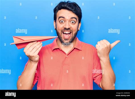 Young Hispanic Man Holding Paper Airplane Pointing Thumb Up To The Side
