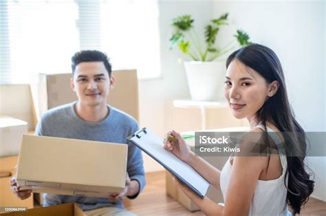 아시아 가족 이사 집 젊은 귀여운 여자는 상자를 들고있는 동안 클립 보드에 종이에 물건을 확인합니다 아내와 남편은 카메라를보고 행복한 얼굴로 미소 택배 입주 및 배송 2명에