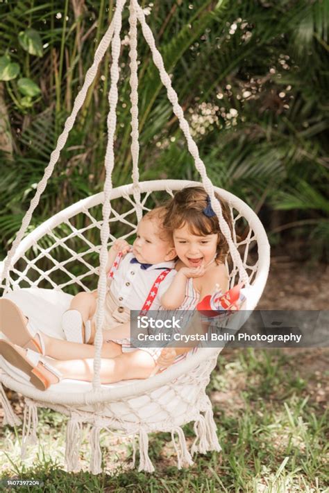 빨간 서스펜더와 파란 활 넥타이를 착용 한 6 개월 된 아기 소년이 3 살짜리 큰 여동생이 빨간색 흰색 및 파란색 줄무늬 드레스를 입고 Boho 교수형 의자에서 스윙하고