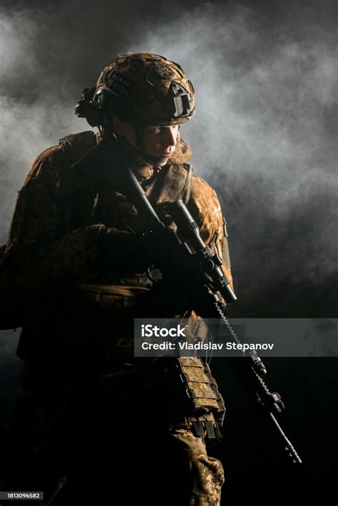군용 위장복 헬멧 마스크를 착용한 육군 병사가 소총을 들고 검은 배경의 연기 속에서 공격 자세로 서서 빨간 점 조준경으로 조준합니다 모션에 대한 스톡 사진 및 기타 이미지
