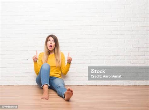 흰색 벽돌 벽을 통해 바닥에 앉아 젊은 성인 여자 놀라게 했 고 올려 손가락으로 가리키는 그리고 팔 놀 랐 어 요 가리키기에