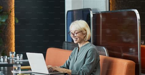 안경을 쓴 쾌활하고 매력적인 성숙한 프리랜서가 주황색 소파가 있는 카페의 테이블에 앉아 노트북으로 타이핑을 하며 새로운 온라인 프로젝트를 진행하고 있다 사진