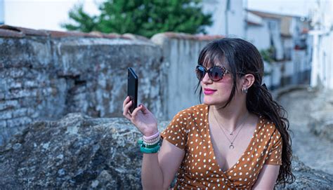 긴 머리와 선글라스를 가진 예쁜 여자는 오후 태양에 돌에 앉아 그녀의 휴대 전화를 본다 14 15 살에 대한 스톡 사진 및 기타 이미지 14 15 살 거리 고름