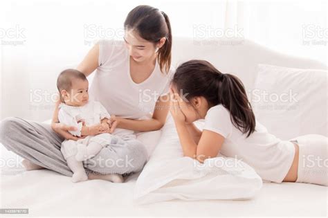나이 든 아시아 자매는 하얀 방에서 침대에서 신생아와 놀고 아름다운 어머니는 아기를 사랑스럽게 안고 사랑과 보살핌으로 보고