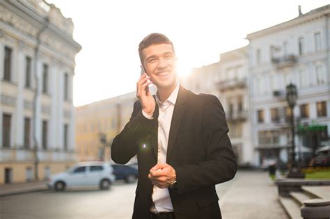 검은 색 재킷과 흰색 셔츠를 입은 젊은 쾌활한 남자가 퍼지 배경에서 아름다운 도시 경관을 가진 휴대 전화로 즐겁게 이야기합니다 20 24세에 대한 스톡 사진 및 기타 이미지