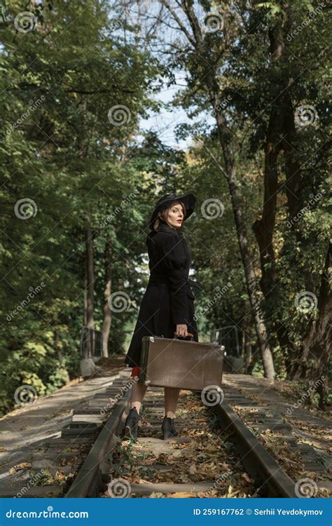 Woman In Black Coat Hat And With Retro Suitcase Wallking On Rails In