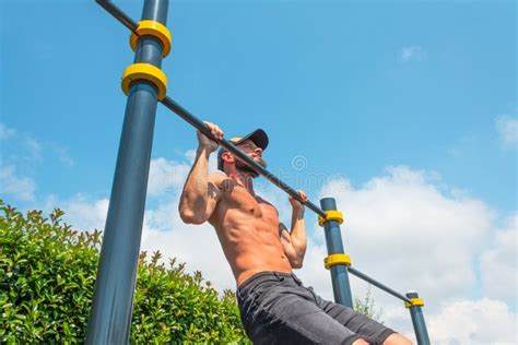 Muscular Man In A Cap Doing Pull Ups On The Horizontal Bar In The Park