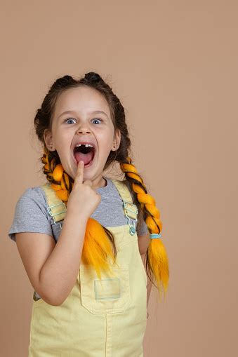 카네칼론 머리띠를 가진 장난기 넘치는 흥분한 어린 소녀의 초상화 베이지 색 배경에 노란색 점프 슈트와 회색 티셔츠를 입은 카메라를 손가락으로 손가락으로 가리키며 찡그린