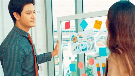 비즈니스 사람들은 사무실의 프로젝트 계획 보드에서 일하고 동료 친구와 대화하여 프로젝트 개발을 분석합니다 그들은 유리 벽에 게시 된 스티커 메모를 사용하여 정리합니다
