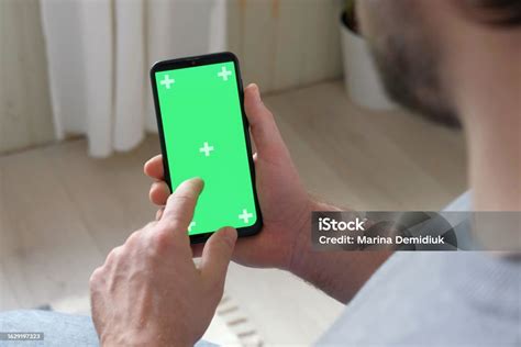 스마트폰 녹색 모형 화면을 손에 들고 집에 앉아 있는 청년 크로마 키 휴대 전화를 사용하는 남성 수직 모드 터치 디스플레이 스 와이프 탭 인터넷 소셜 미디어 서핑 후면에