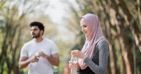 젊은 여성은 녹색 공원에 서 있는 동안 물이 담긴 병을 들고 옷을 입고 히잡을 들고 있습니다 옆으로 보고 있는 그녀의 무슬림 잘생긴 남편의 흐릿한 배경 사진