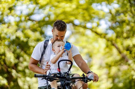 건강한 아침 루틴과 활동적인 가족 유아는 자전거 활동 중에 상쾌한 추위를 마십니다 소년은 자전거 바구니에 앉아 파란색 병에서 물을 마시고 한 남자가 머리에 키스합니다 2명에