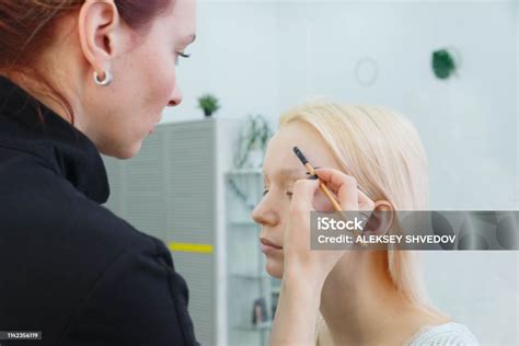 메이크업을 만드는 과정 메이크업 아티스트 모델 면에서 브러시로 작업 합니다 뷰티 살롱 인테리어에 젊은 금발 여자의 초상화입니다 피부에 톤을 적용 고객에 대한 스톡 사진 및