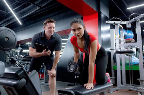 백인 남성 트레이너 지도와 아시아 여성은 피트니스 체육관에서 동기 부여와 헌신으로 덤벨을 들어 올리고 코치 지원 힘 및 건강을 통해 근육질의 손과 팔을 위한 여성 운동 2