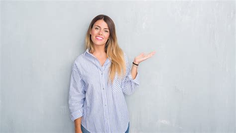 쾌활 한 제시 하 고 카메라를 보고 손바닥으로 가리키는 웃 패션 비즈니스 옷을 입고 회색 그런 지 벽에 젊은 성인 여자