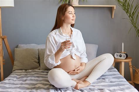 흰 옷을 입고 배꼽을 드러낸 매력적인 꿈꾸는 임산부는 밝은 침실에서 침대에 앉아 배를 만지고 건강을 돌보며 신선한 물을 마신다