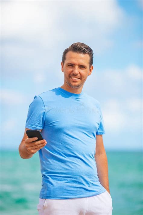 Young Man Holding Mobile Phone In His Hand While Walking On The Beach