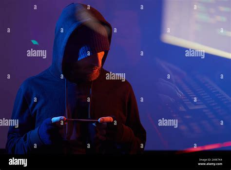 Bearded Computer Hacker In Black Hoody Shirt Holding Smart Phone And Working Online In Dark Room