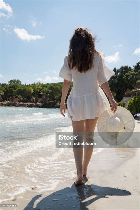 하얀 태양 드레스를 입고 모자를 손에 들고 여름에 모래 사장을 걷는 아름다운 젊은 여성 끈 원피스에 대한 스톡 사진 및 기타 이미지 끈 원피스 후면 걷기 Istock