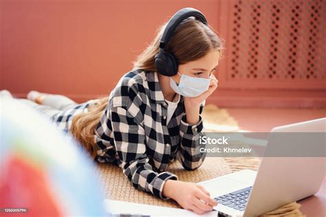 보호 의료 마스크를 착용한 소녀는 노트북으로 바닥에 앉아 온라인 응용 프로그램을 통해 교사와 혼아합니다 원격 학습 온라인 교육