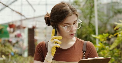 안경을 쓴 젊은 여성 꽃집은 전화로 고객과 이야기하면서 문서에 따라 꽃의 가용성을 확인합니다 사진 Unsplash의 온실 이미지