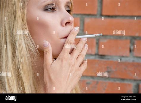 Beautiful Young Blonde Girl Smokes A White Cigarette On A Brick Wall