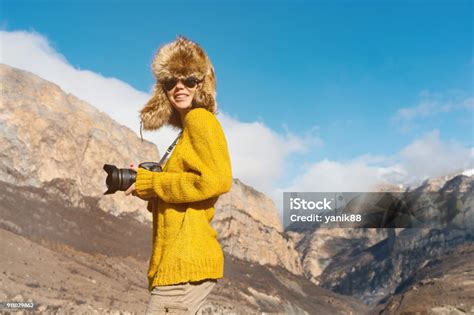 선글라스와 큰 모피 모자와 노란색 니트 스웨터에 여자 사진 작가 서 계곡에서 높은 바위의 배경에 대 한 Dslr 카메라와 함께 그녀의 손에 가죽에 대한 스톡 사진 및 기타