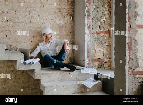 Smiling Architect Sitting With Laptop And Blueprints On Steps At Site