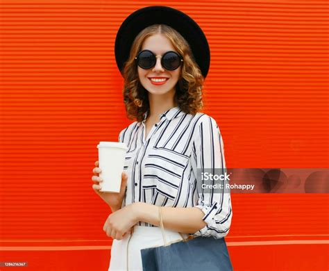 커피 컵과 흰색 줄무늬 셔츠를 입은 쇼핑백 빨간 배경 위에 검은 둥근 모자를 쓴 아름다운 웃는 여성의 초상화 여름에 대한 스톡 사진 및 기타 이미지 Istock