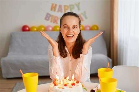 혼자 테이블에 앉아 케이크와 촛불로 생일을 축하하고 카메라를 보고 손을 들어 긍정적 인 감정을 표현하는 젊은 성인 매력적인