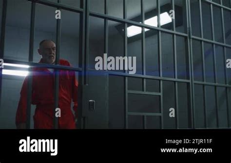 Elderly Criminal In Orange Uniform Stands Near Barred Window Leans On