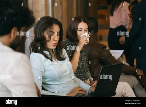 Lovely Girl Taking A Sip Of Coffee And Looking At Her Colleague Lap Top